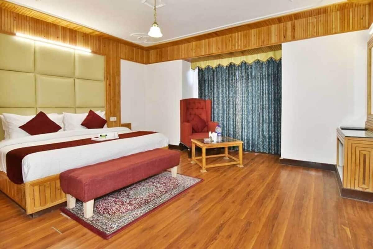 Quarto de hotel com piso de madeira, cama com cabeceira, poltrona vermelha, mesa pequena e janela com cortinas.