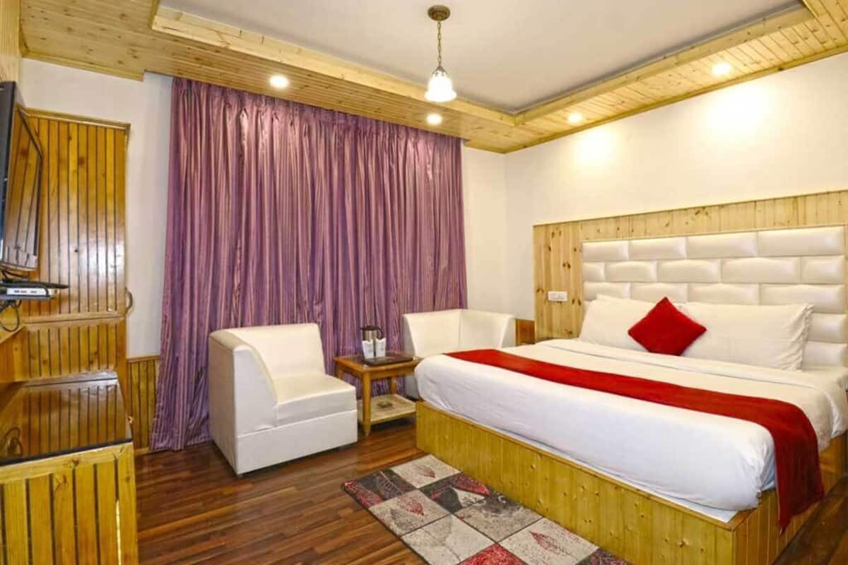 Quarto com cabeceira de madeira, cama com roupa de cama branca e um travesseiro vermelho, poltrona branca, mesa de cabeceira de madeira e cortina roxa.