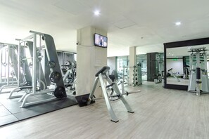 Uma academia com diversos aparelhos de ginástica, uma televisão fixada na parede e um espelho grande refletindo os equipamentos.