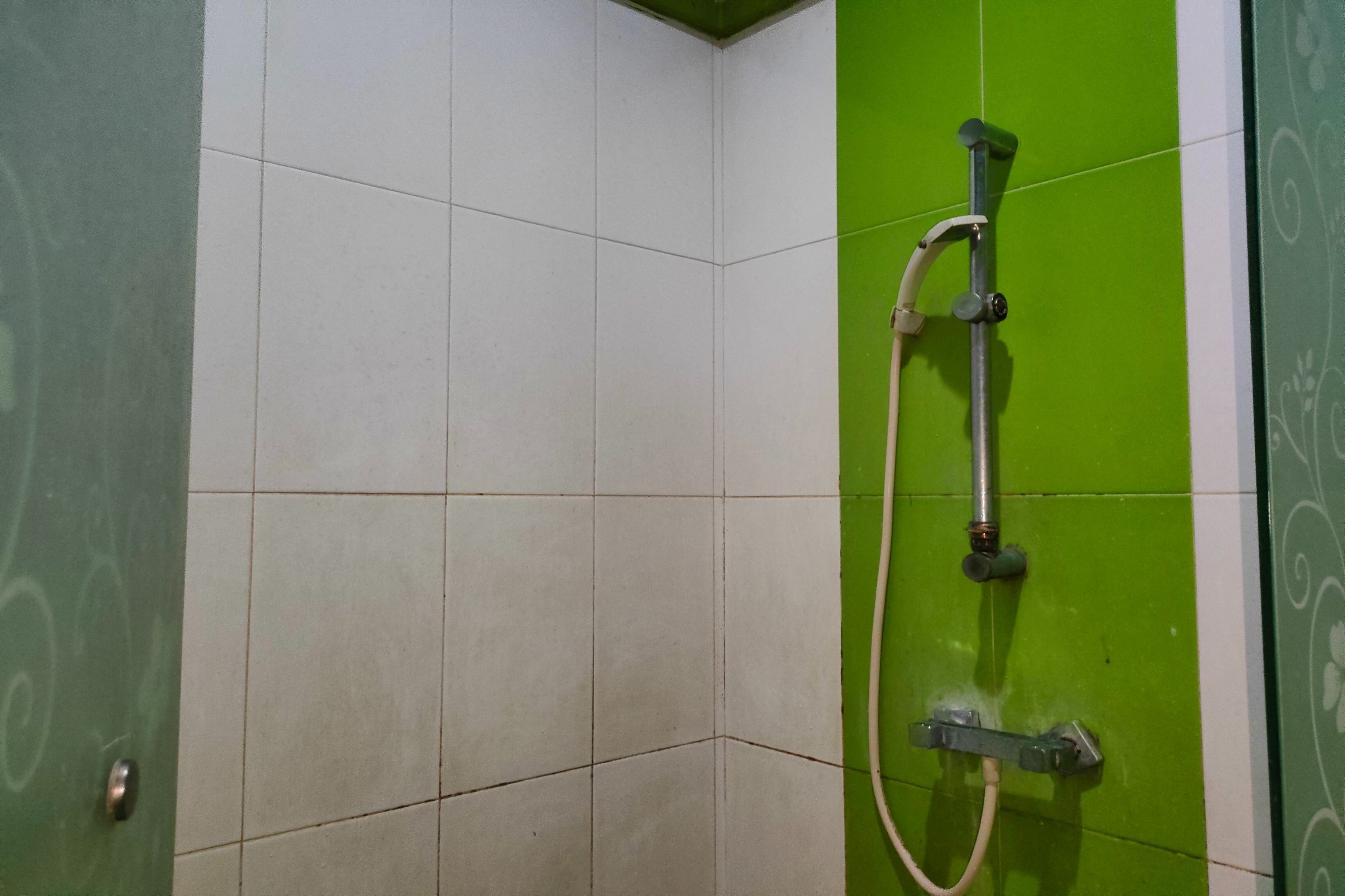 Canto de um banheiro com azulejos brancos e verdes, um chuveiro e um cano aparente.