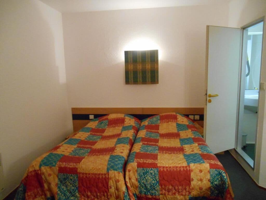 Quarto com duas camas, uma luminária na parede e porta de acesso a um banheiro.