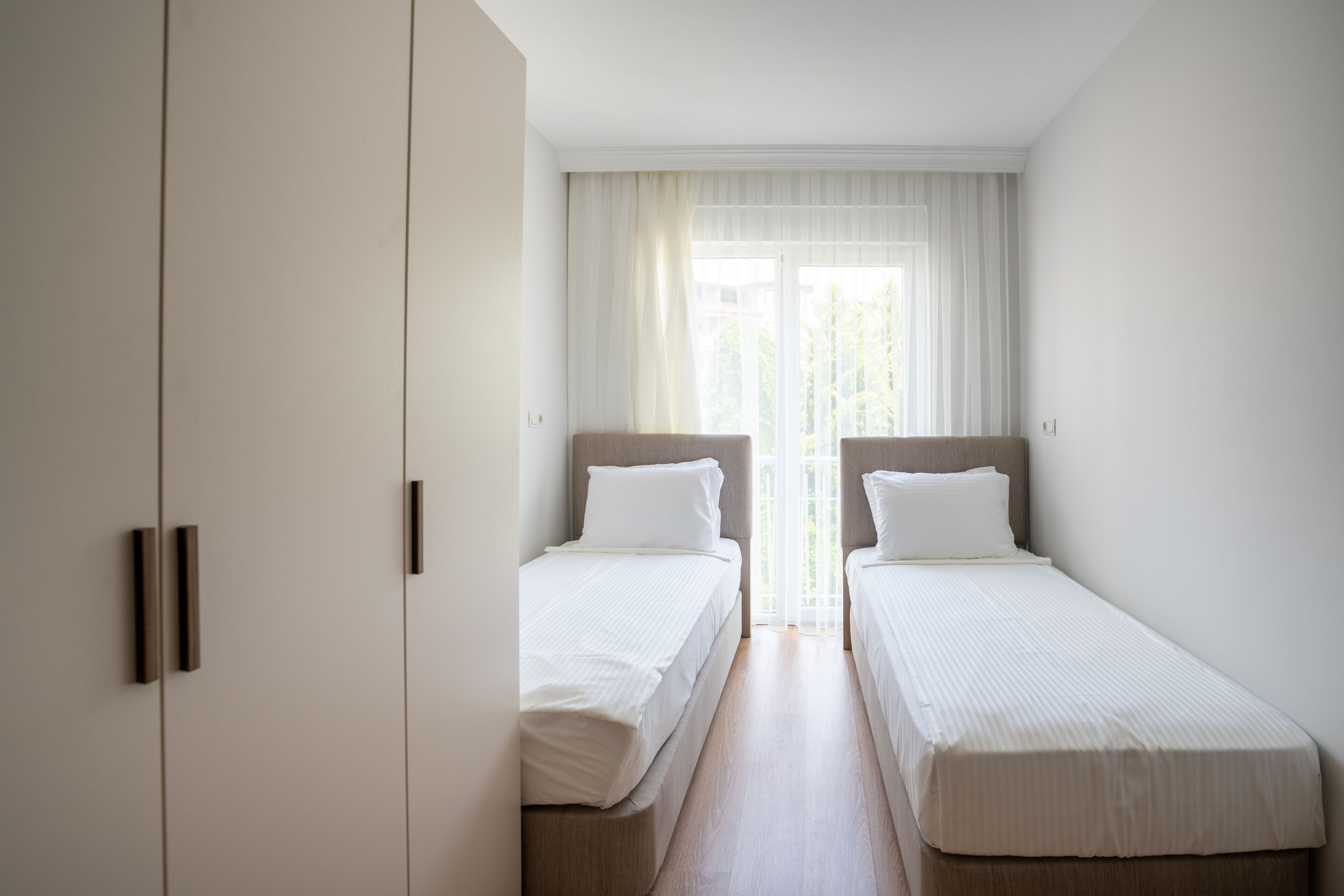 Quarto com duas camas de solteiro, um guarda-roupa e uma janela grande com cortinas leves.