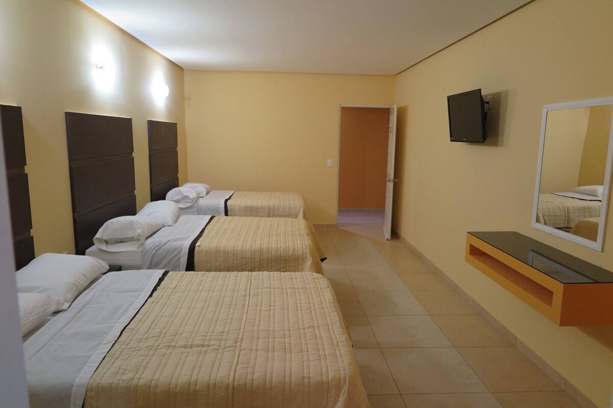 Quarto de hotel com três camas, uma TV fixada na parede e uma pequena escrivaninha com espelho.