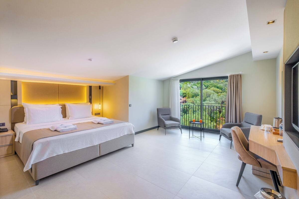 Quarto de hotel moderno com uma cama grande, varanda com assentos e vista para o verde.