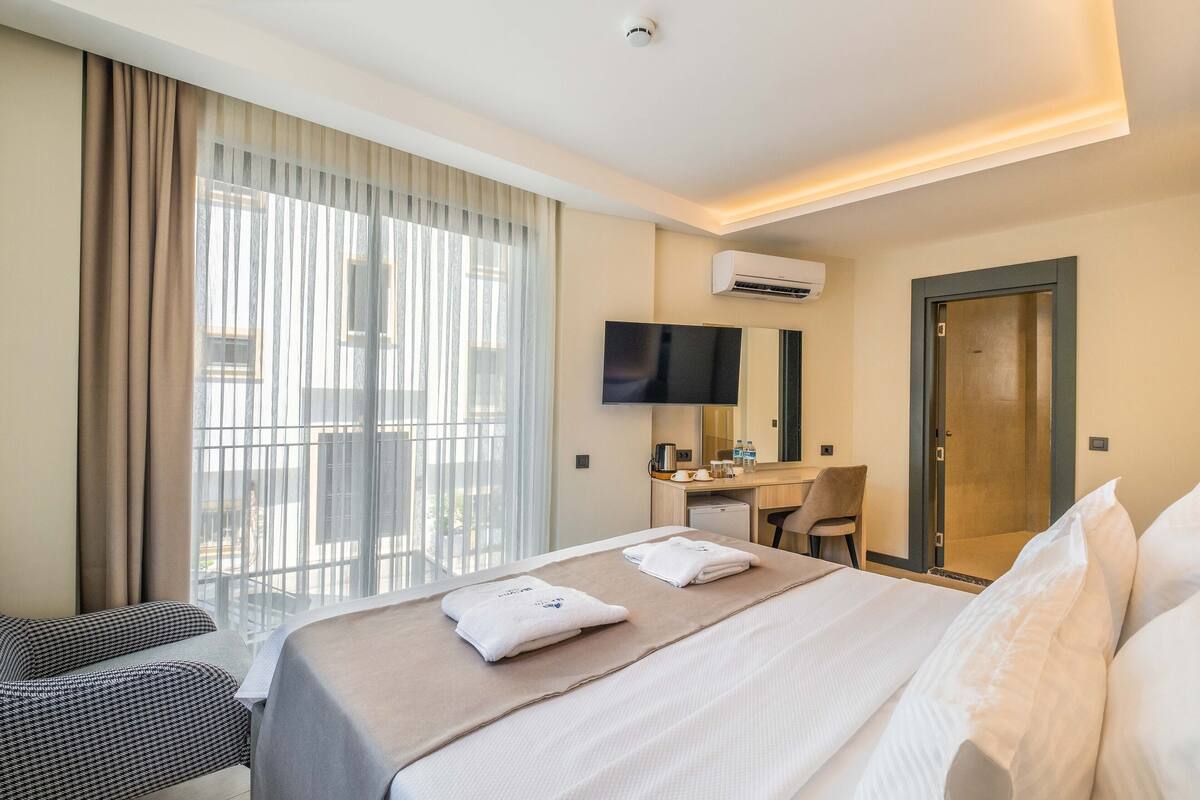 Quarto de hotel com cama, escrivaninha, cadeira, televisão e janela com cortinas.