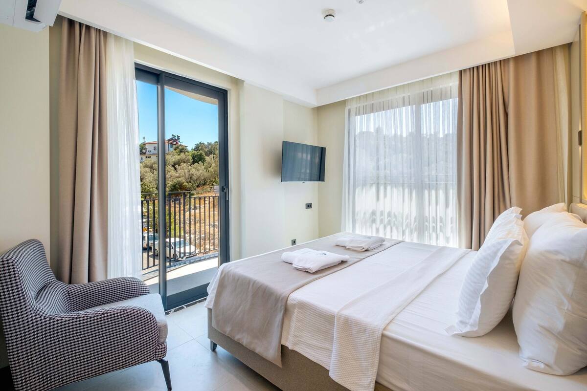 Quarto de hotel com cama, uma cadeira, televisão e varanda com vista para árvores e edifícios.