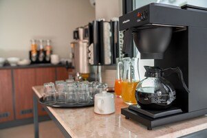 Máquina de café em bancada de cozinha, com um suporte de copos, uma caneca branca e um jarro de suco de laranja.