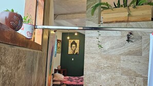 Quarto com teto de madeira, parede verde e cama com colcha estampada. Há uma prateleira com plantas e um porta-retrato na parede.
