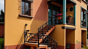 Uma casa de dois andares com um pátio pavimentado em pedra, uma escadaria com grades de metal e uma varanda com plantas em vasos.