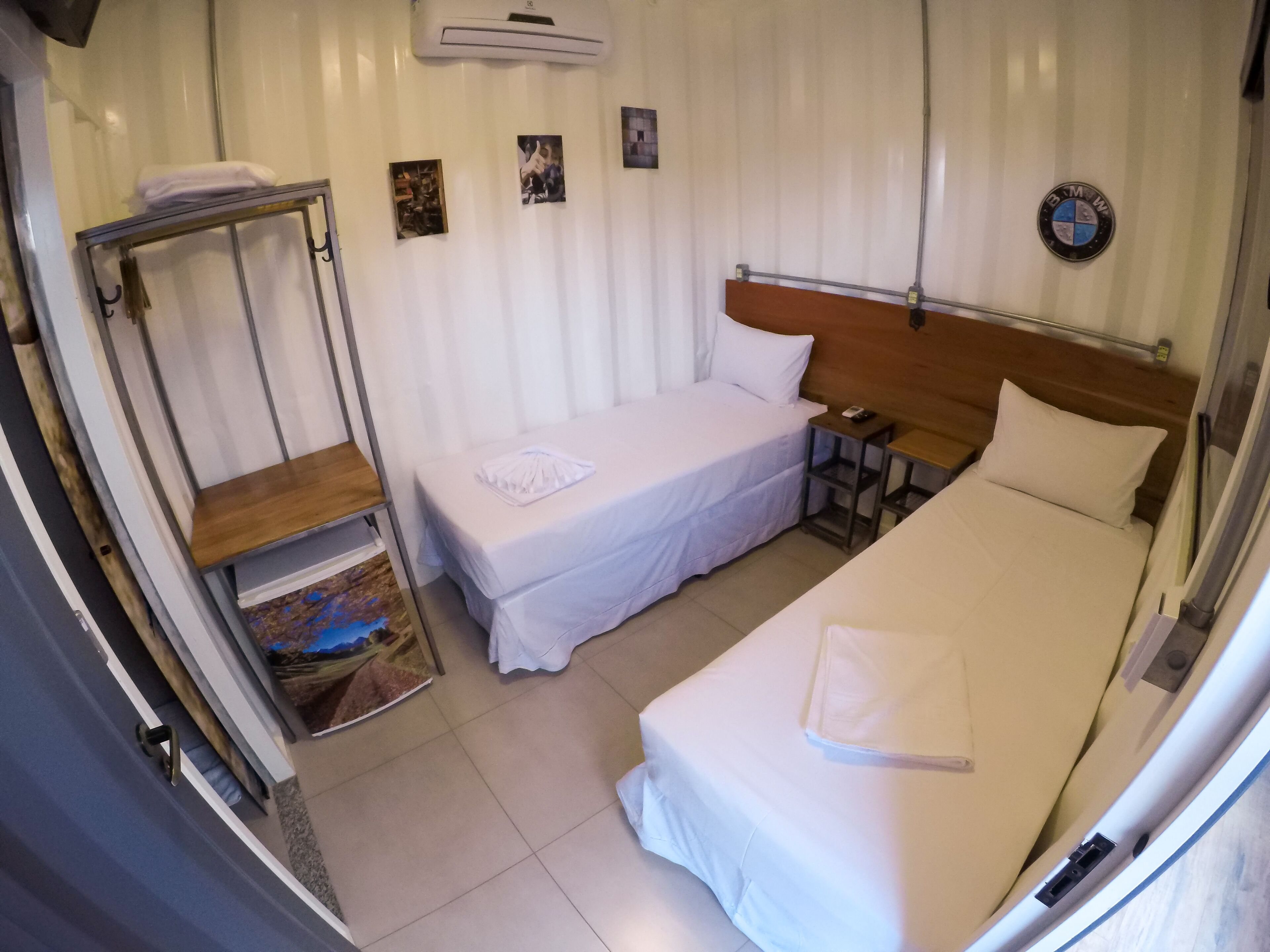Quarto de hotel com duas camas, uma cabeceira de madeira, uma mesinha pequena e uma estante com toalhas.