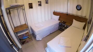 Quarto de hotel com duas camas, uma cabeceira de madeira, uma mesinha pequena e uma estante com toalhas.