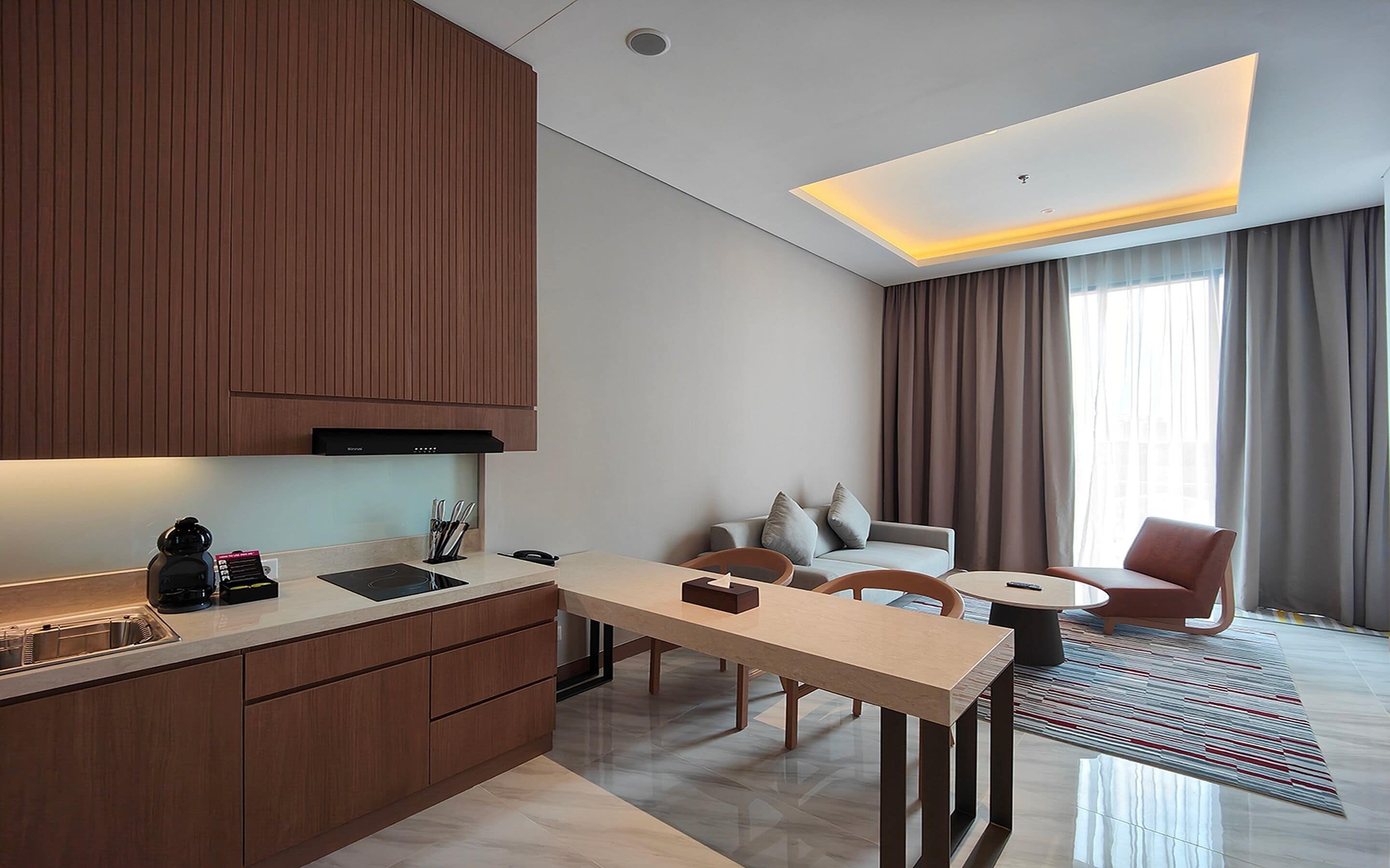 Quarto de hotel moderno com cozinha compacta, espaço para refeições e área de estar com sofá e poltronas.