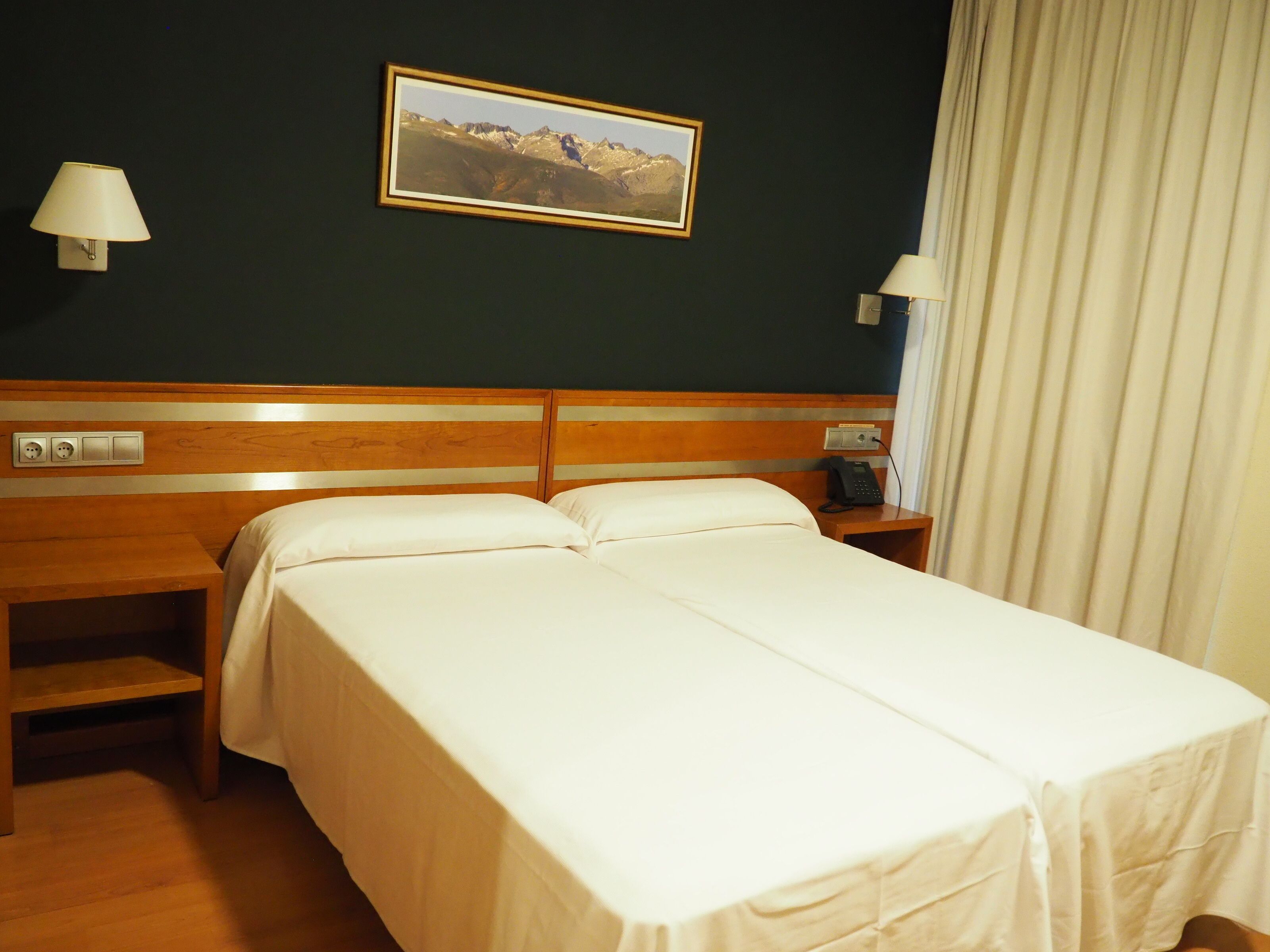 Quarto de hotel com cama de casal, duas mesas de cabeceira, luminária na parede, telefone e pintura de paisagem montanhosa.