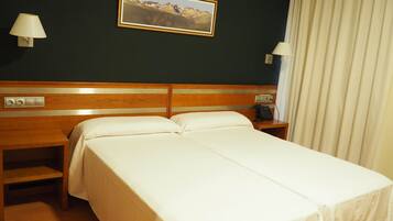 Quarto de hotel com cama de casal, duas mesas de cabeceira, luminária na parede, telefone e pintura de paisagem montanhosa.