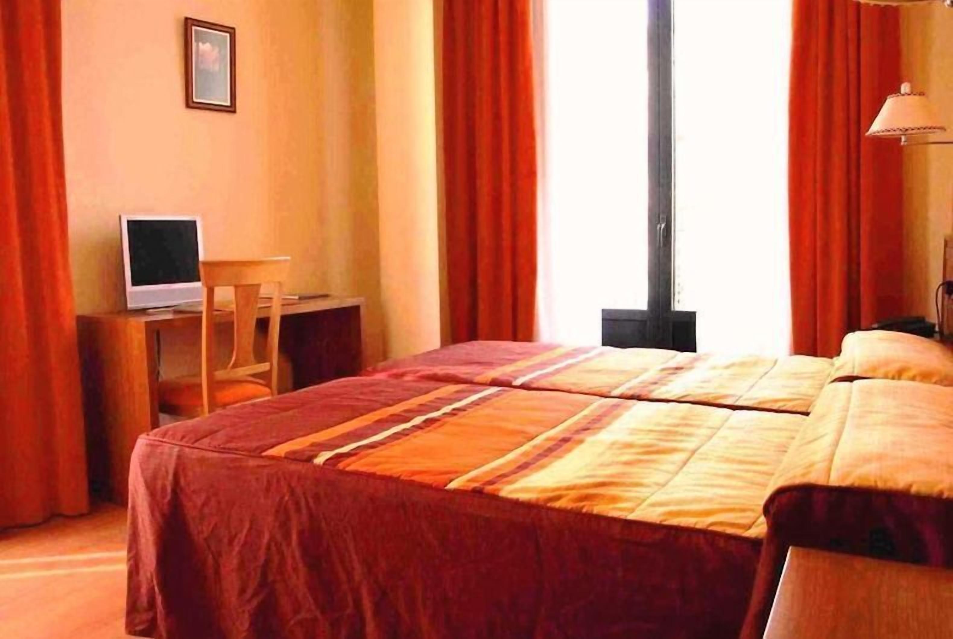 Quarto de hotel com cama, escrivaninha e computador, e janela com cortinas.