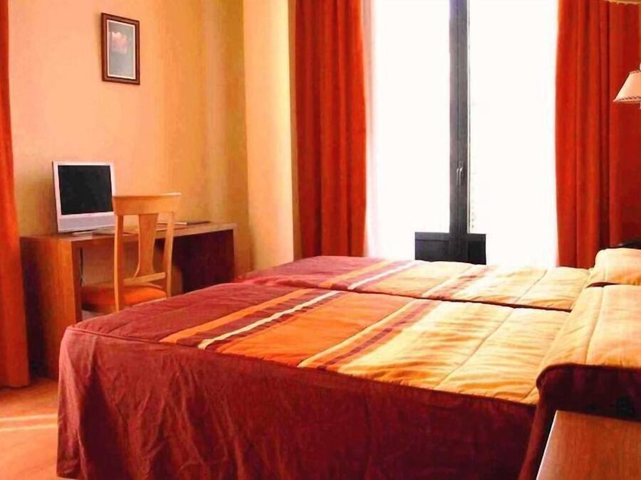 Quarto de hotel com cama, escrivaninha e computador, e janela com cortinas.
