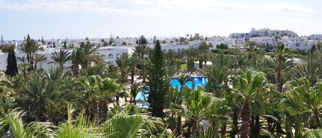 Um resort com piscina cercada por palmeiras, edifícios brancos ao fundo e céu claro.