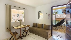 Um quarto pequeno e acolhedor com sofá, mesa de jantar e cadeiras, janela com cortinas e uma rede lá fora.