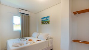 Cama bem arrumada com lençóis e toalhas brancas, um quadro na parede e uma prateleira de madeira com um objeto pequeno.