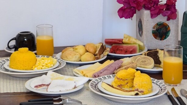 Uma mesa de pequeno-almoço com vários pratos, incluindo ovos, torradas e fruta. Há um vaso com flores e uma máquina de café na mesa.