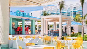 Um resort com área de piscina, cadeiras e mesas amarelas, um bar chamado 'License to Chill' e um edifício com varandas.