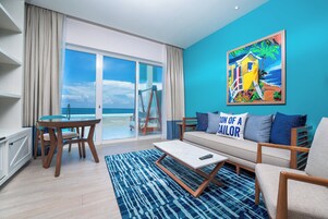 Uma sala de estar moderna com uma grande porta de vidro deslizante que dá para uma varanda, um quadro de uma casa na praia, e um sofá com uma almofada com a inscrição "SON OF A SAILOR".