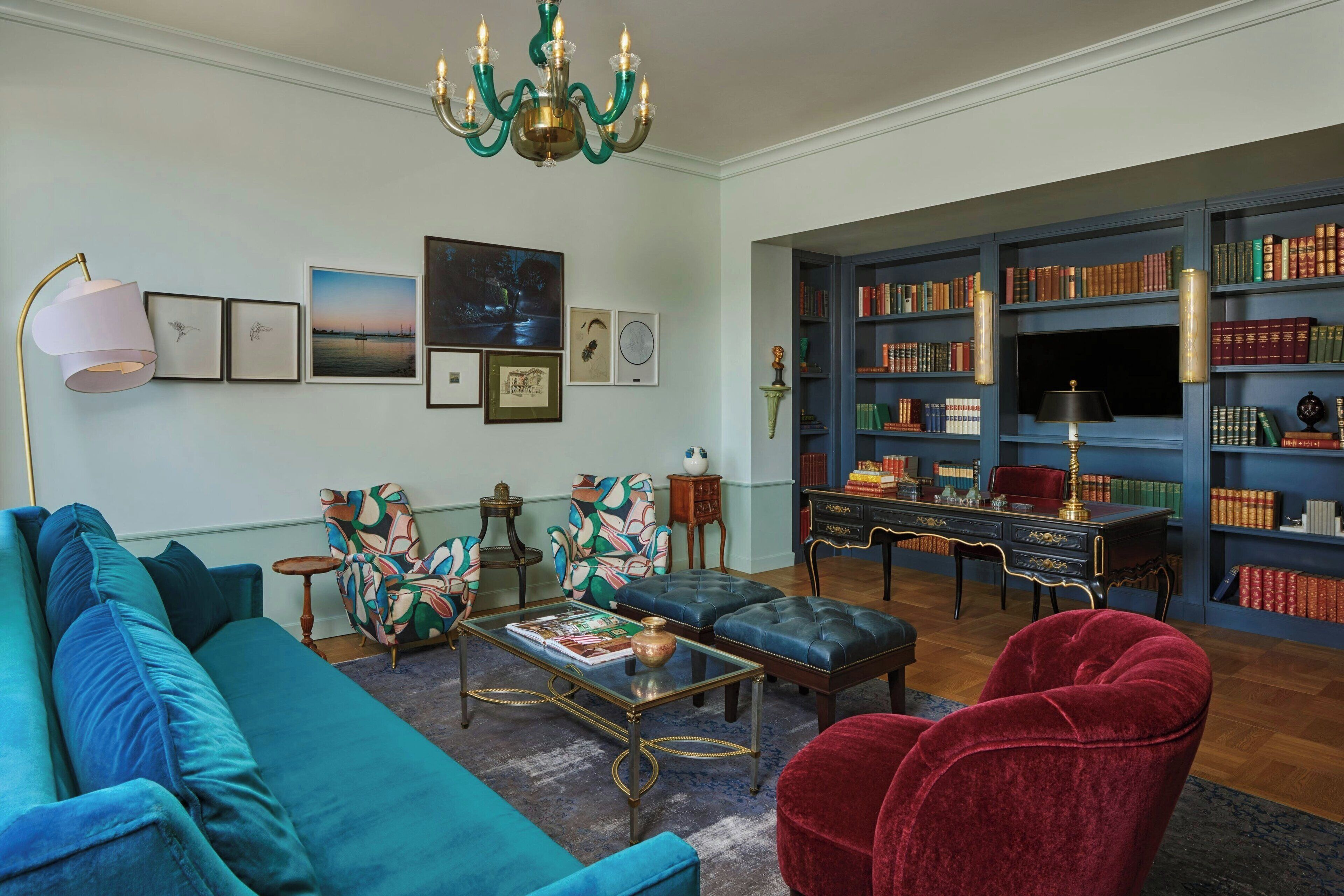 Uma sala de estar com um sofá verde-água, uma poltrona de veludo vermelho, uma mesa de centro e uma estante repleta de livros.