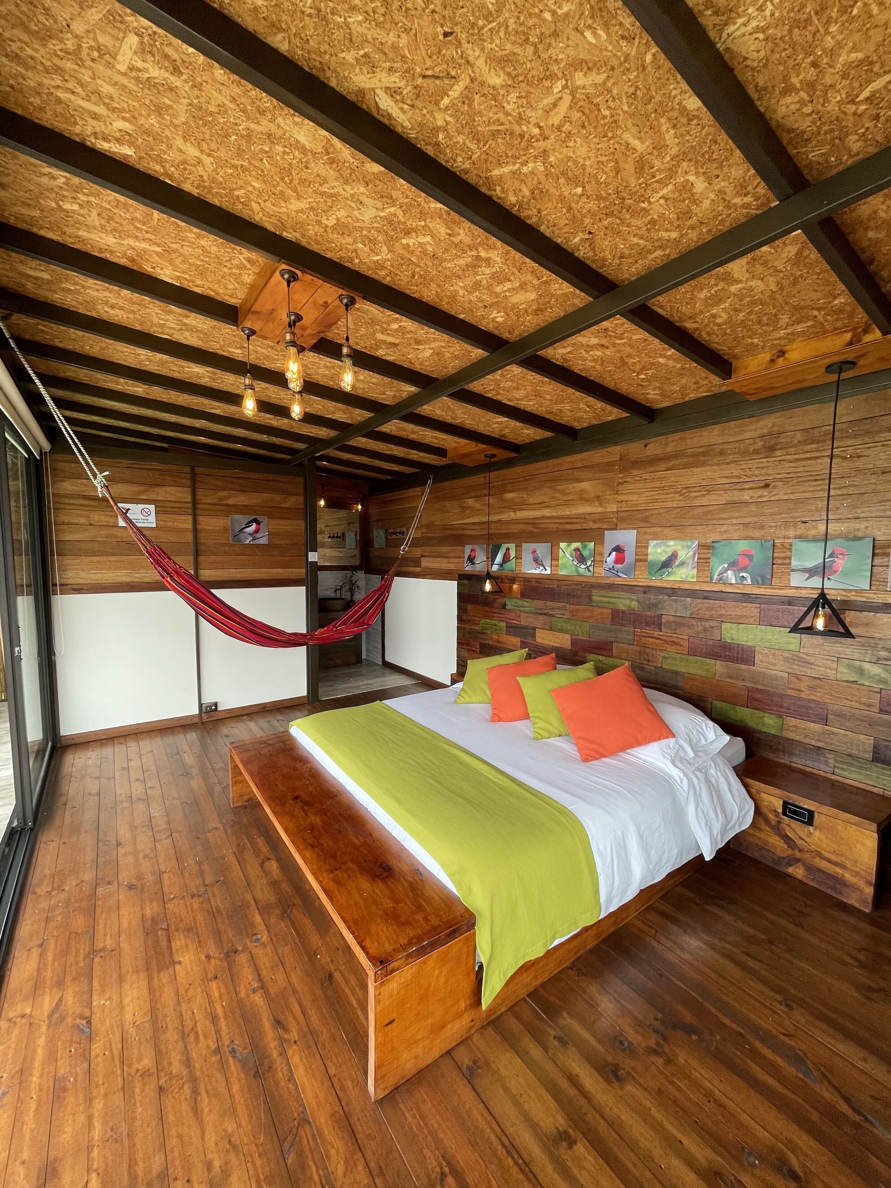 Quarto com piso de madeira, cama com colcha verde e laranja, uma rede e parede com pinturas de pássaros.