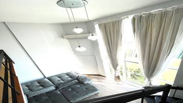 Uma sala de estar moderna com um sofá cinzento, luminárias pendentes e janelas amplas com cortinas.