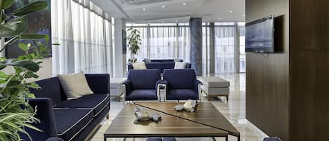Um lobby moderno de hotel com área de estar, janelas amplas e uma TV de tela plana na parede.