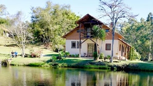 Uma casa de dois andares com varanda, localizada ao lado de um lago tranquilo, rodeada por árvores e uma área gramada.