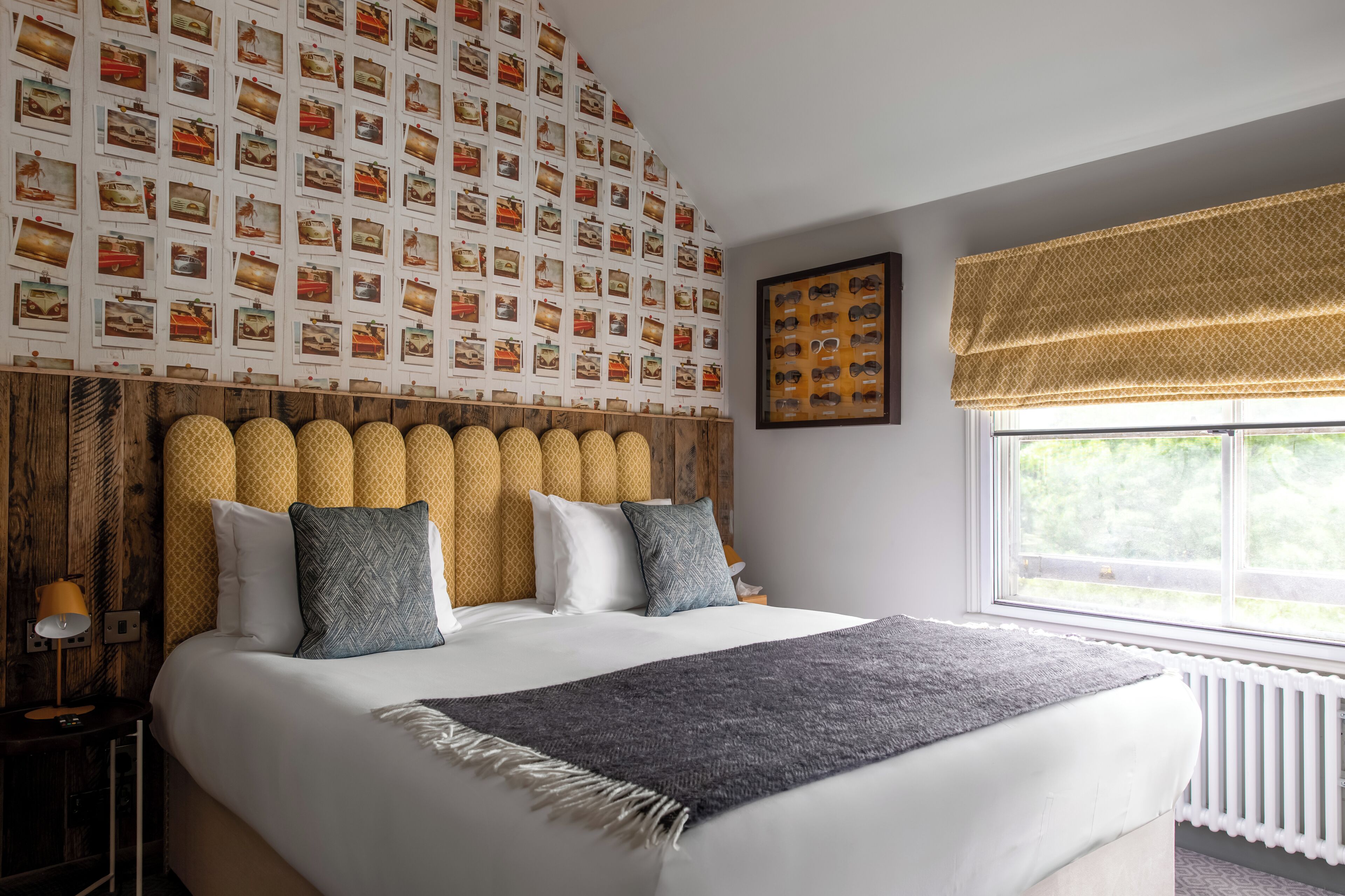 Um quarto bem arrumado com uma cama grande, parede estampada, janela com cortina e uma moldura com óculos de sol.