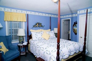 Quarto com cama de dossel, sofá azul, mesa de cabeceira com abajur e decoração floral na parede.