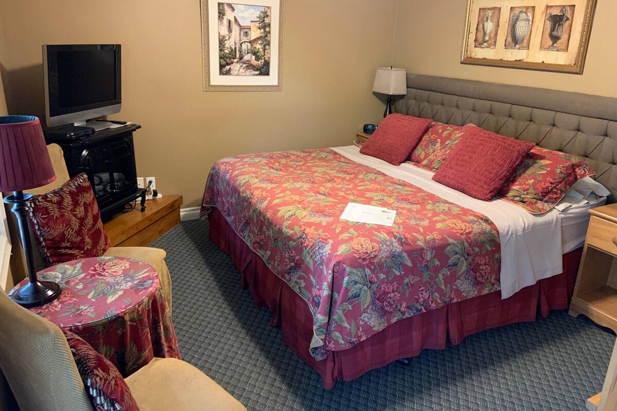 Quarto de hotel com uma cama grande, televisão, lareira, mesa pequena com abajur e obras de arte nas paredes.