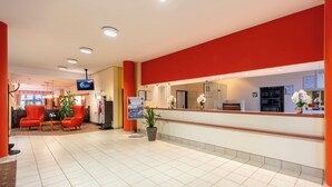 Um saguão de hotel com balcão de recepção, duas poltronas laranja, uma mesa de centro, um vaso de plantas e uma tela de televisão.