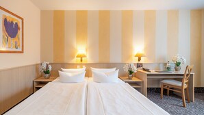 Quarto de hotel com duas camas, uma escrivaninha, uma cadeira e uma luminária na parede.