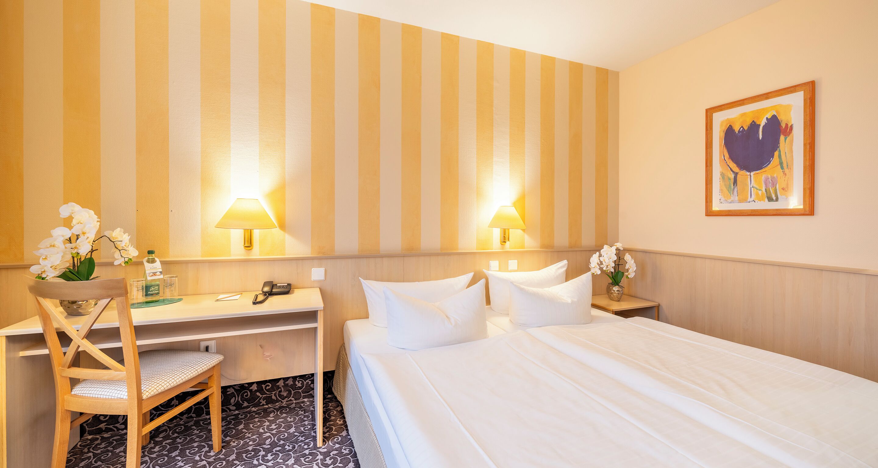 Quarto de hotel com cama, escrivaninha, cadeira, luminárias e um quadro na parede.