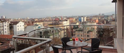 Varanda com mesa e cadeiras, vista para um horizonte urbano com diversos edifícios e uma cadeia montanhosa ao longe.