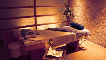 Uma sala de massagem com uma cama, uma mesinha com velas, uma cadeira e um vaso com planta.