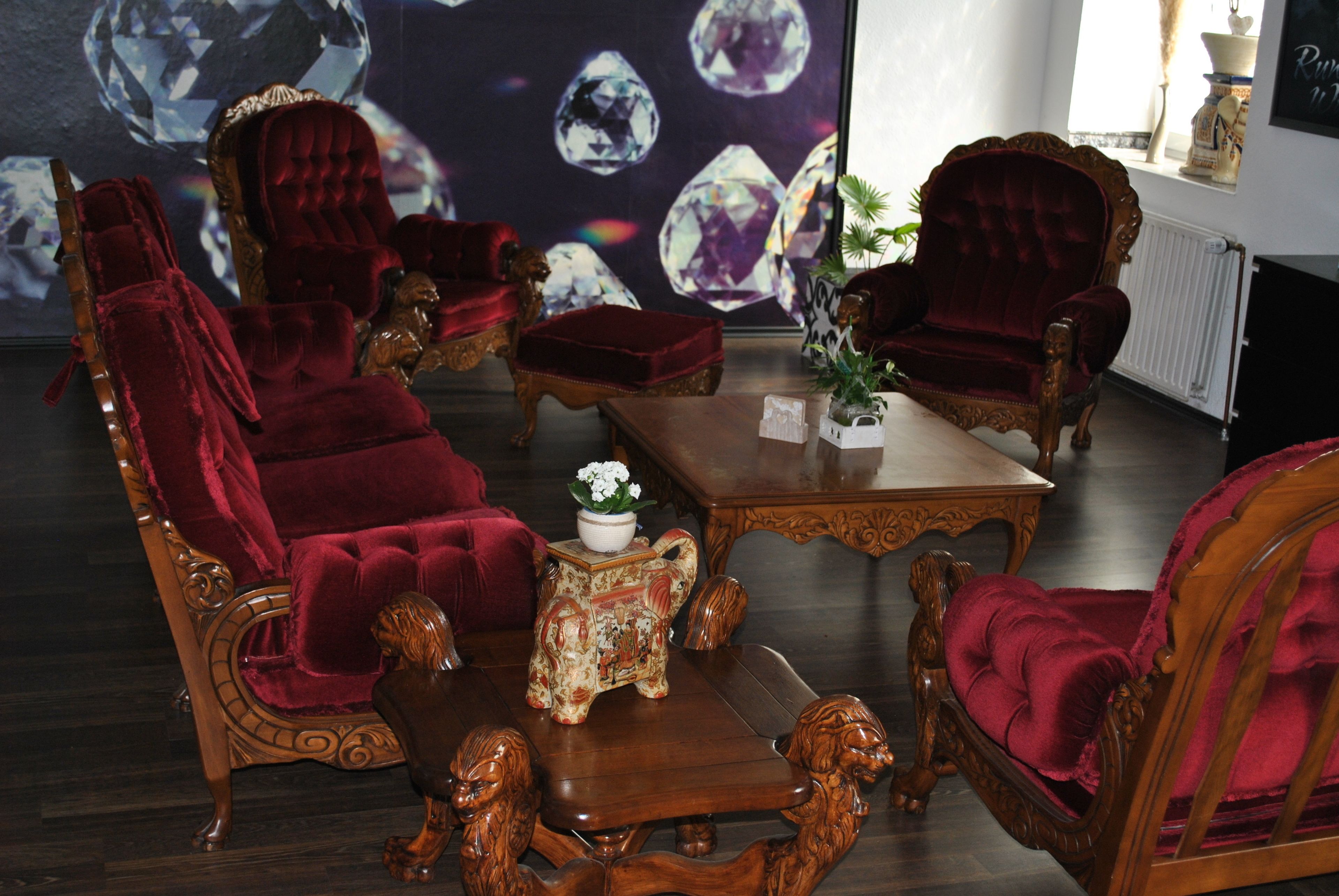 Quarto com mesa de centro de madeira, duas poltronas de veludo vermelho e um banquinho combinado. Também se vê uma mesinha lateral com uma planta.