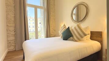 Quarto com uma cama grande, um espelho redondo e vista para edifícios pela janela.