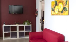 Uma sala de estar com um sofá vermelho, uma mesa de console branca e uma televisão de tela plana montada acima dela, além de um quadro com limões na parede.