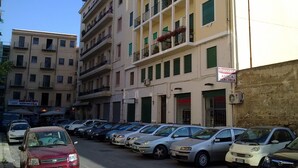 Uma fileira de carros estacionados ao longo de uma rua ladeada por edifícios altos, alguns com sacadas e placas indicando estabelecimentos comerciais.