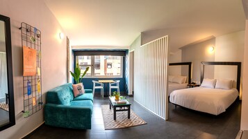 Quarto de hotel compacto com uma cama, um sofá, uma mesinha e uma janela.