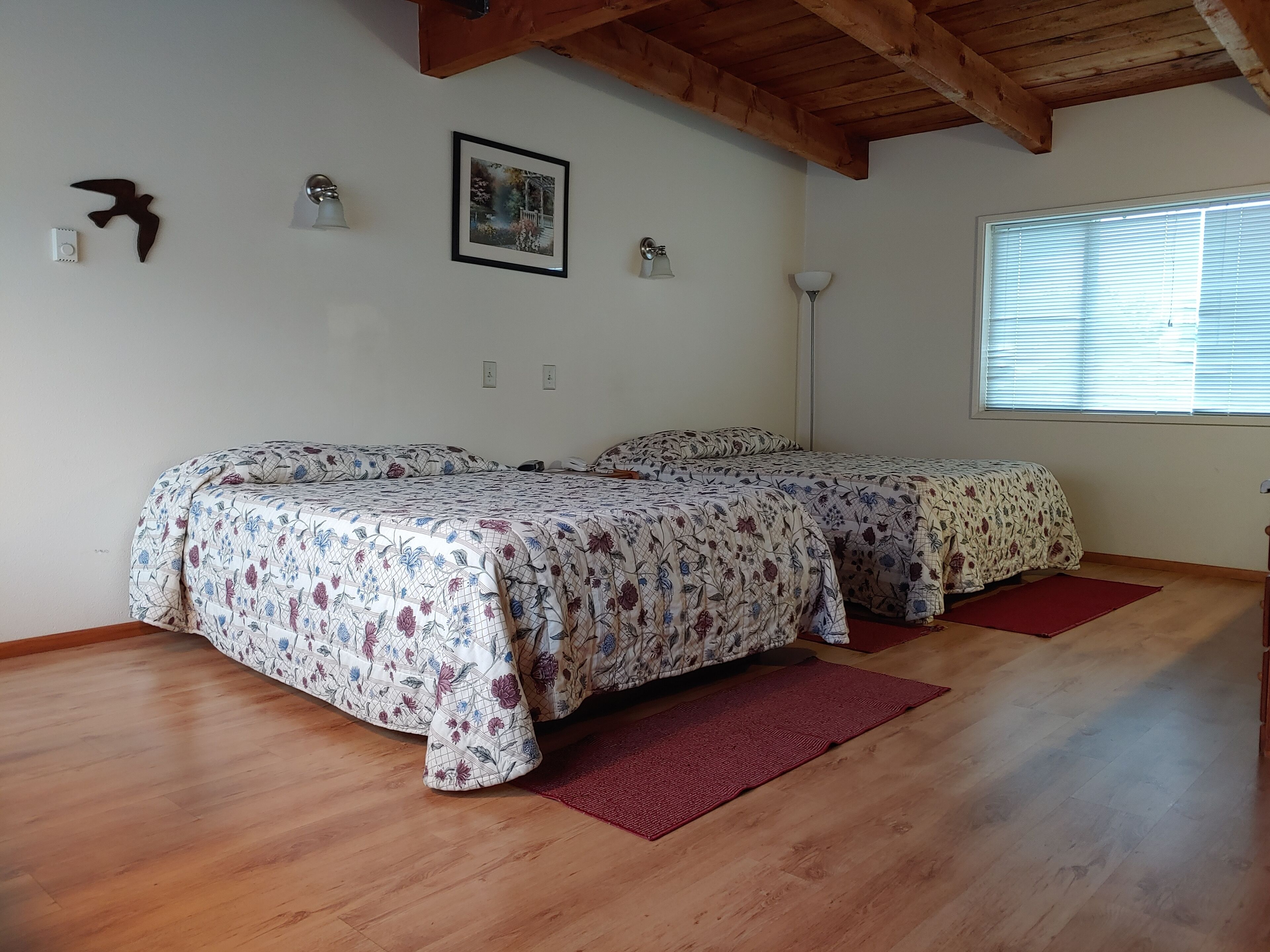 Quarto com duas camas, piso de madeira, tapete vermelho, janela com persianas e teto com vigas aparentes.