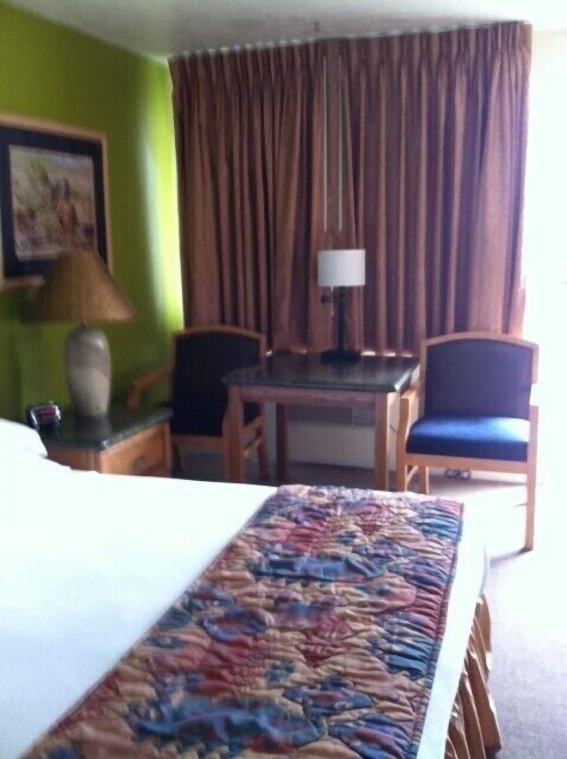 Quarto de hotel com uma cama, uma escrivaninha, duas cadeiras, um abajur e um quadro na parede.