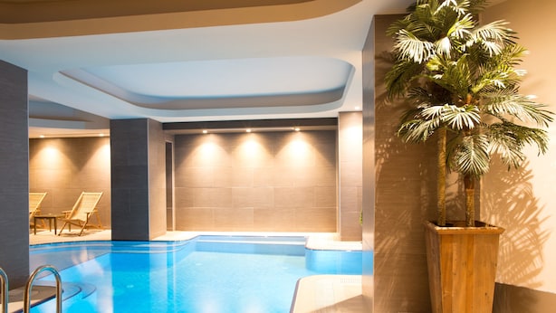 Piscina interna com água azul cristalina, cercada por paredes revestidas de azulejos e um vaso de madeira com uma palmeira.