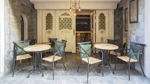 Uma área de refeições com quatro mesas redondas e cadeiras, cada uma com assento de fibra trançada e estrutura de metal. As paredes são decoradas com desenhos padronados complexos, e um candelabro pendurado no teto.