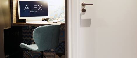 Quarto de hotel com uma escrivaninha, uma cadeira e um computador. O quarto possui ladrilhos estampados e uma porta.