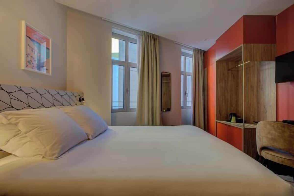 Quarto de hotel com cama, cadeira, televisão e janela com cortinas.
